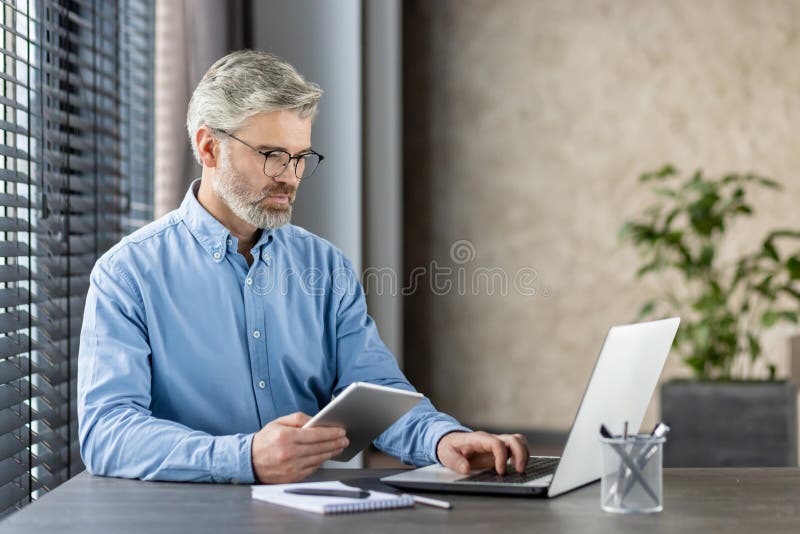 Mature Office Worker Focused on a Task while Using a Laptop and Tablet ...