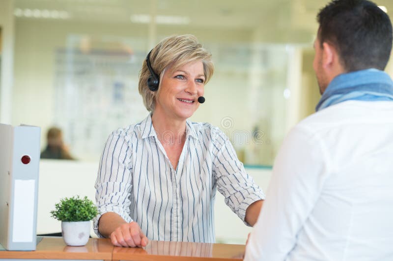 Mature Receptionist Wearing Headset Speaking To Customer at Counter ...