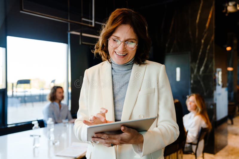 Mature Woman in Jacket Smiling and Using Tablet during Offline Meeting ...