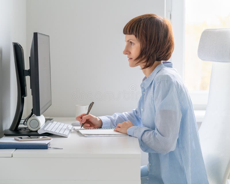 Mature Woman Looking At Computer Screen And Writing Information In ...