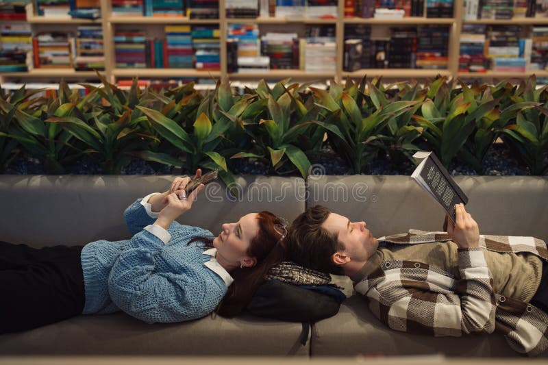 Two People Reading and Using Smartphone on Couch in Library Stock Photo ...