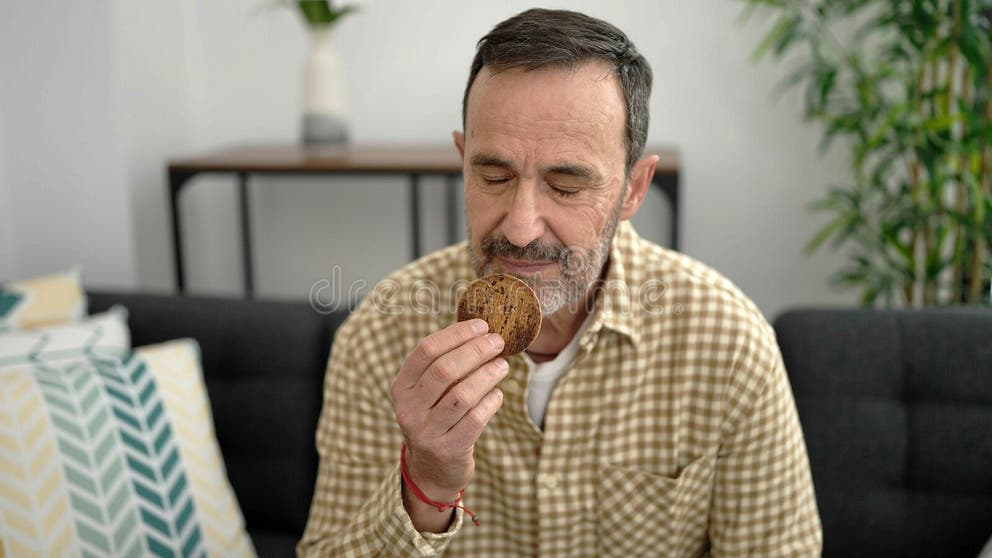 Middle Age Man Eating Cookie Sitting on Sofa at Home Stock Photo ...