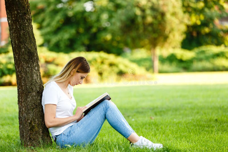 Modern Student Sitting in Park Under the Tree and Reading the Bo Stock ...