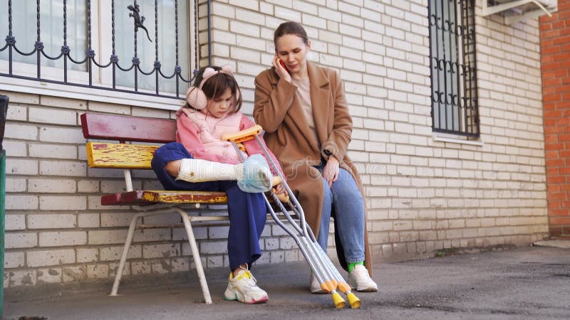 Mom Talking On Phone Daughter With Crutches On Bench In Yard Stock