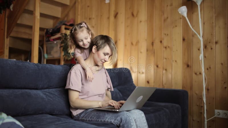 Mother Is Trying To Work On Laptop With Daughter Cuddling Her In Living