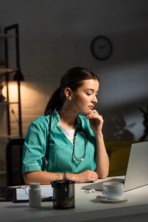 Nurse in Uniform Sitting at Table and Looking at Laptop during Night ...