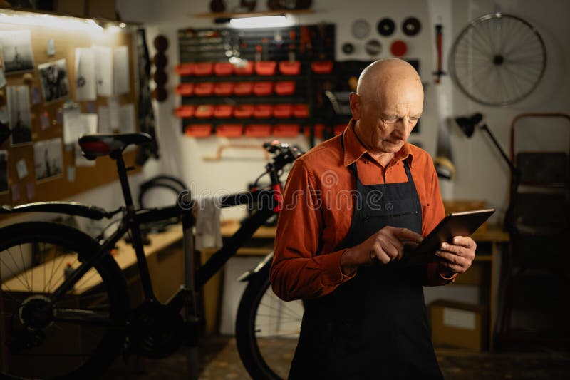 Old Mechanic Using Digital Tablet Repairing a Bicycle in a Workshop or ...