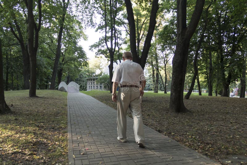 An Old, Stooped Man Walks Sullenly and Alone Along the Path in the Park ...