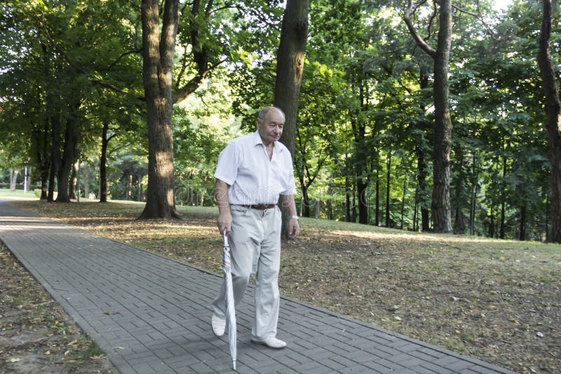 An Old, Stooped Man Walks Sullenly and Alone Along the Path in the Park ...