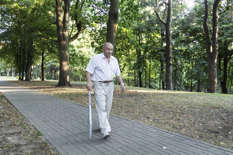 An Old, Stooped Man Walks Sullenly and Alone Along the Path in the Park ...