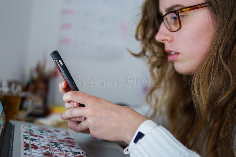 Photograph of a Girl Looking at the Mobile in Front of Her Computer at ...