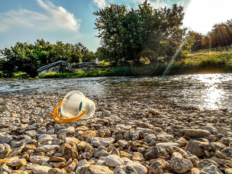 Plastic Bucket Lying on a Rocky Riverbank with Trees and Sunlight in ...