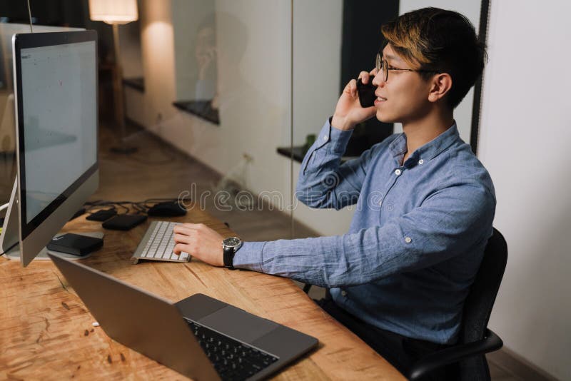 Pleased Asian Guy Talking on Cellphone while Working with Computers ...