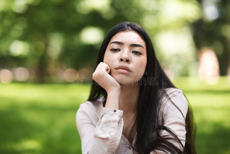 Pensive Asian Girl Sitting On Lawn In Park And Looking Away Closeup