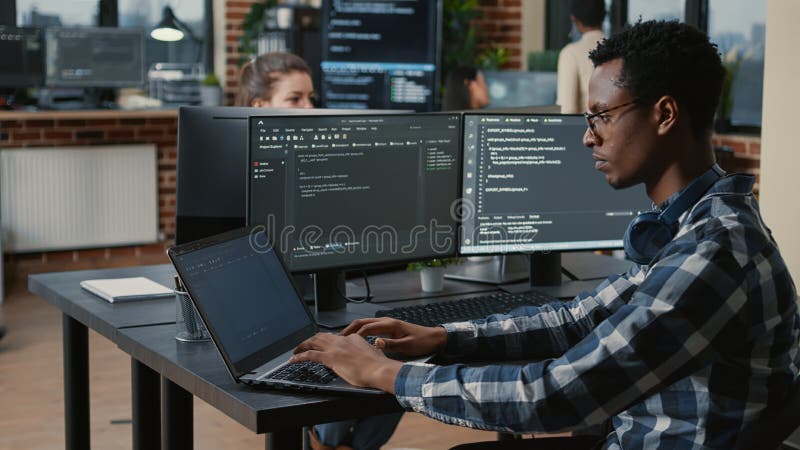 Portrait Of Tired Programer Rubbing Eyes While Typing On Laptop Sitting At Desk With Multiple