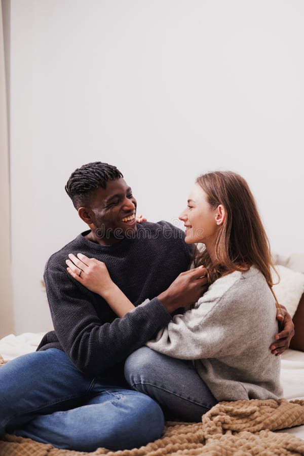 Positive Young Multiethnic Couple Hugging and Talking on Bed at Home ...