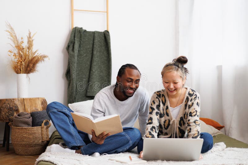 Positive Young Multiethnic Couple Using Laptop and Reading Book on Bed ...
