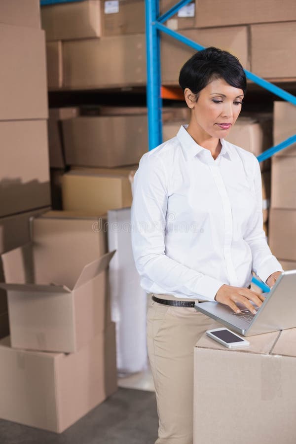 Female Warehouse Clerk Leaning on Cardboard Boxes Checking Laptop and Smartphone in Storage Area ...