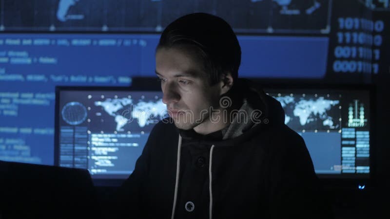 Young Geek Hacker Programmer is Working on Computer in Cyber Security Center Filled with Display ...