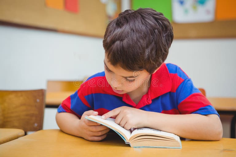 Boy Sitting at Wooden Desk in Classroom Reading Paperback Book Using ...