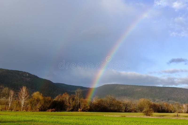 Rainbow on Dark Rain Clouds Over Green Fields with Trees and Mountains ... Rainbow on Dark Rain Clouds Over Green Fields with Trees and Mountains ...