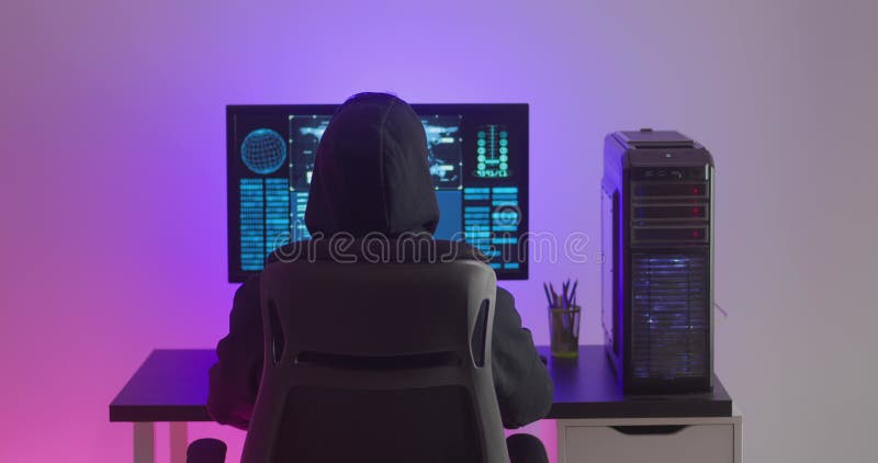 Rear View of Anonymous Hacker in Hood Working at Computer in a Data Center at Night. Man ...