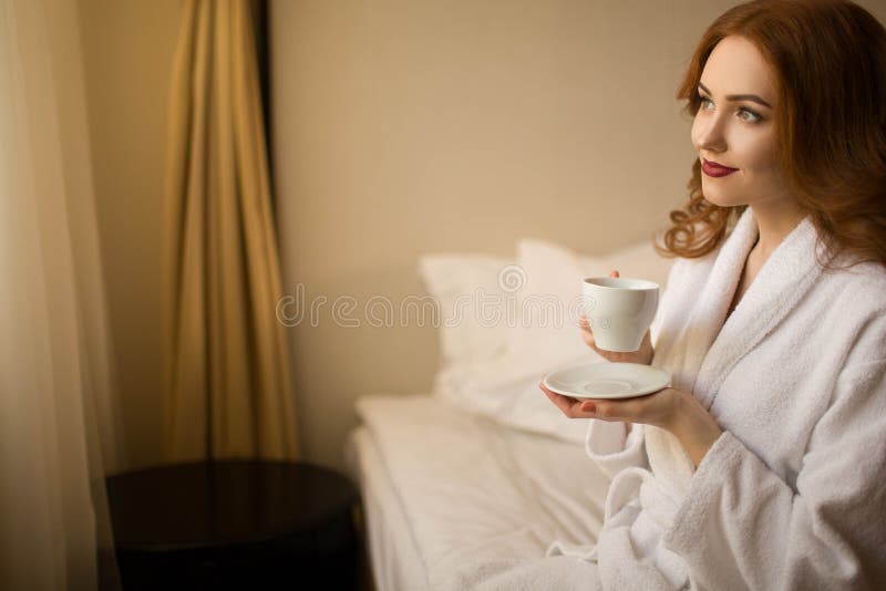 Red-haired Girl in a White Robe with a Cup of Coffee Sitting on a Bed ...