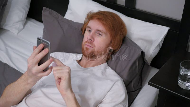 Redhead Man Lying in Bed Using Internet on Smartphone Stock Photo ...
