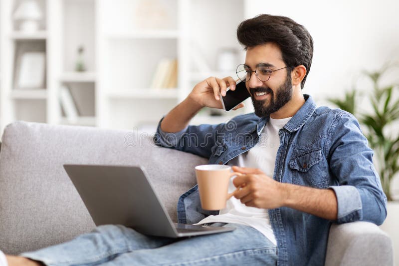 Remote Work. Smiling Indian Man Using Laptop and Cellphone at Home ...
