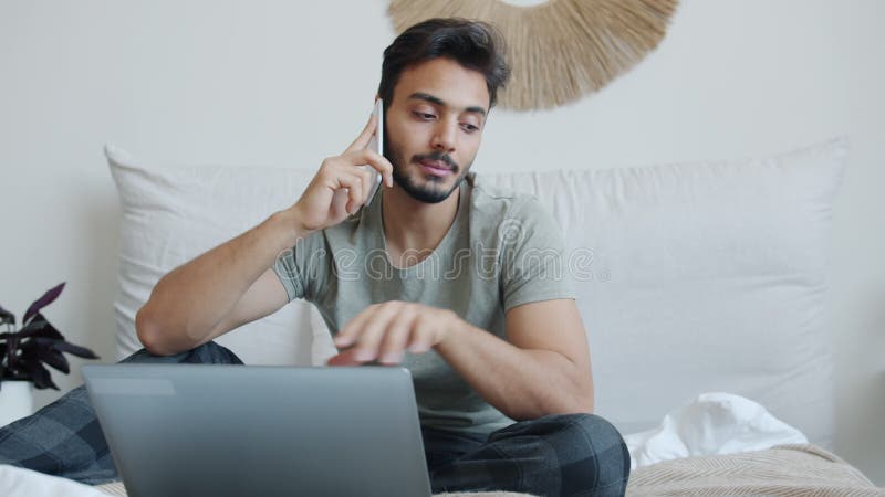 Remote Worker Calling on Mobile Phone and Using Laptop Working in Bed ...
