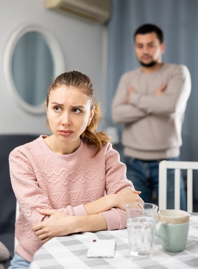 Sad Woman Sitting At Table At Home Husband Quarreling With Her Stock