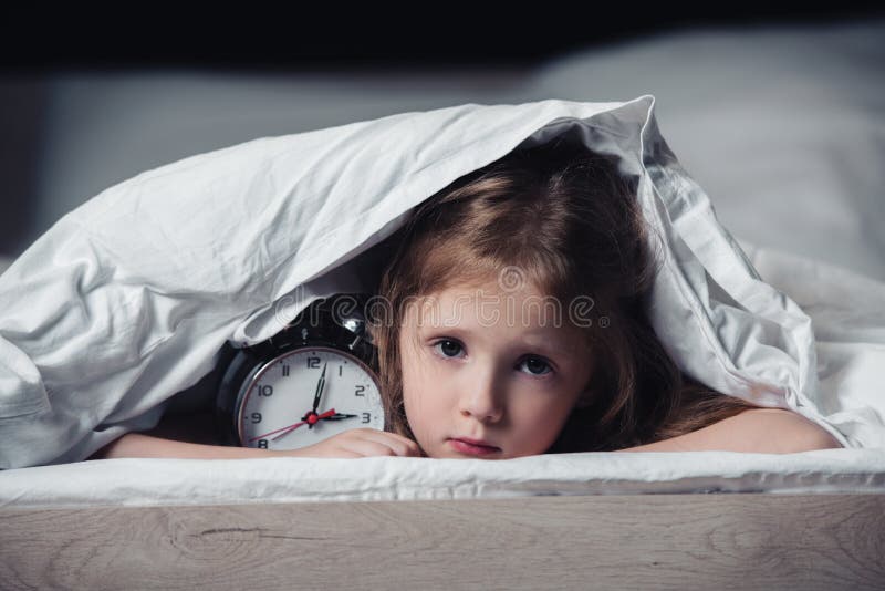 Scared Kid Hiding Under Blanket with Alarm Clock and Looking at Camera ...