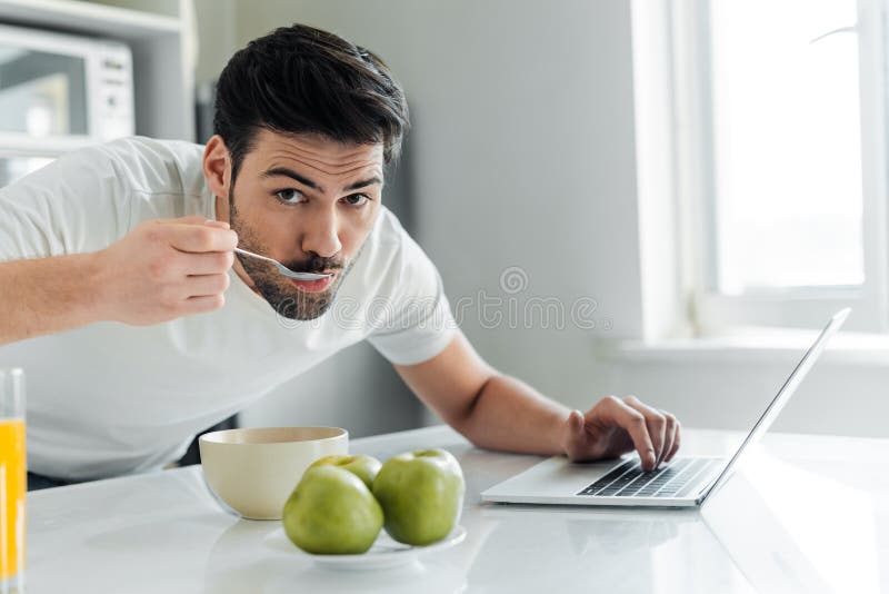 Focus of Man Looking at Camera while Eating Breakfast and Using Laptop ...