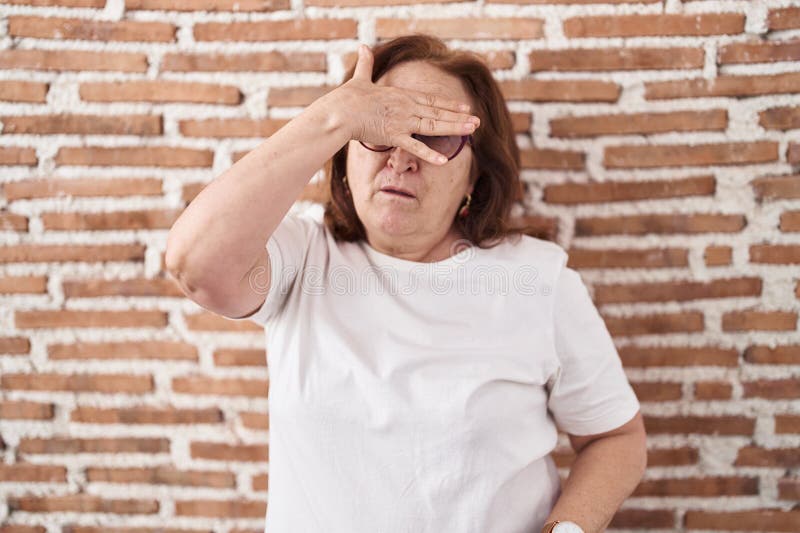 Senior Woman With Glasses Standing Over Bricks Wall Covering Eyes With
