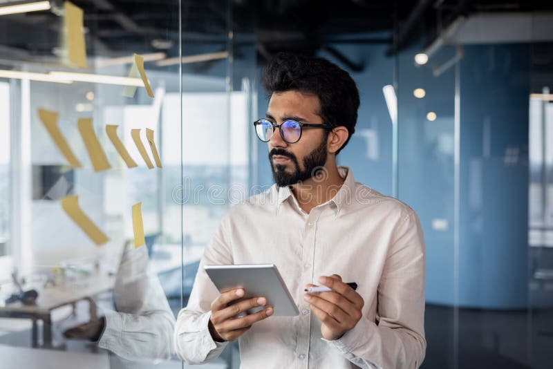 Serious and Focused Young Indian Man Working in Office with Tablet, Standing Near Glass Board ...