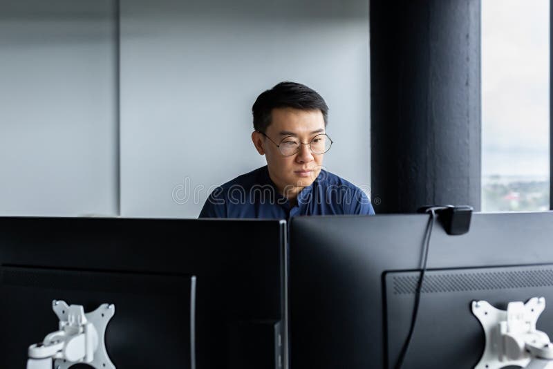 A Serious Young Asian Man Works at Computers in a Modern Office. he ...
