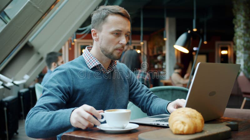Serious Young Man Using Laptop Working and Drinking Coffee in Modern ...