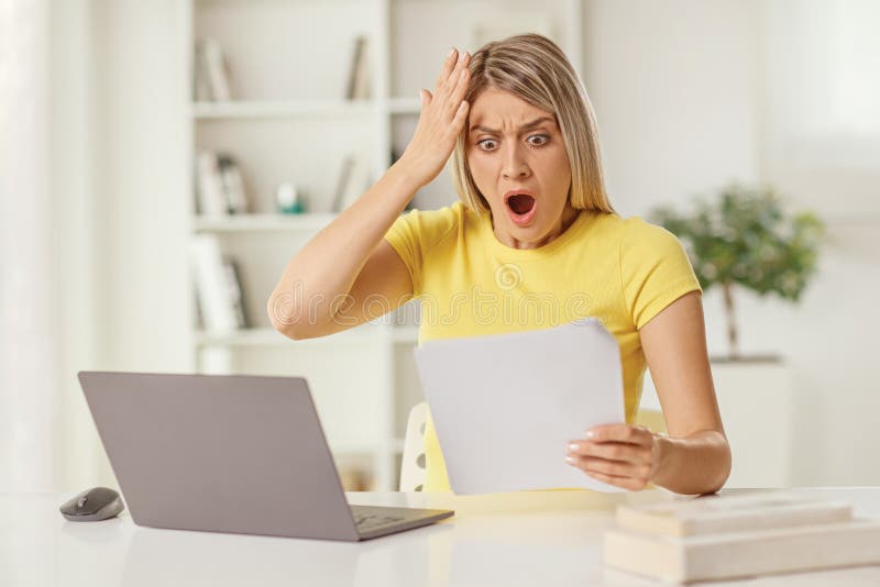 Shocked Young Woman Looking at a Document and Sitting at a Laptop ...