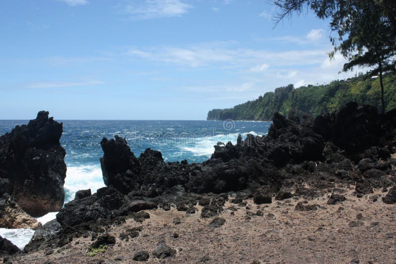 Shoreline Of Large Jagged Black Lava Rock And Boulders And Cliffs Covered In Vegetation In