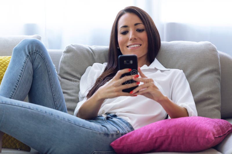 Pretty Young Woman Using Her Mobile Phone While Sitting On Sofa At Home