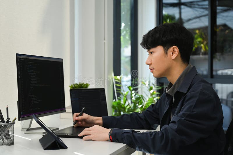 Side View Of Asian Male Web Developer Is Writing Program Code Software On Computer Screens Stock