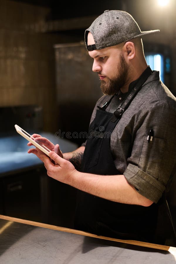 Side View of Young Serious Cook in Uniform Scrolling through Online ...