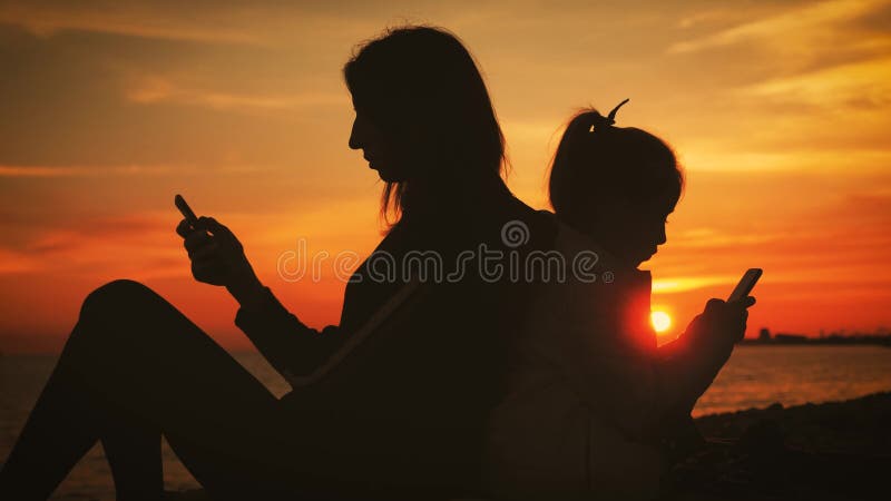 Silhouettes Mother And Daughter Use Mobile Phone On The Beach At Sunset