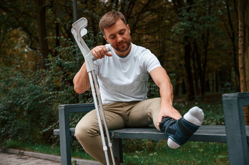 Sitting, Touching the Leg. Man with Crutches is in the Park Outdoors ...