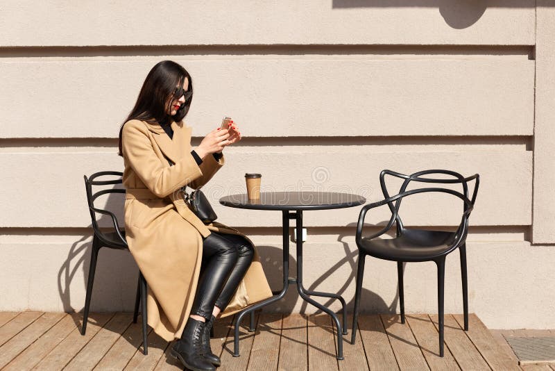 Slender Black Haired Female Sits At Table Outdoors Having Cup Of Hot