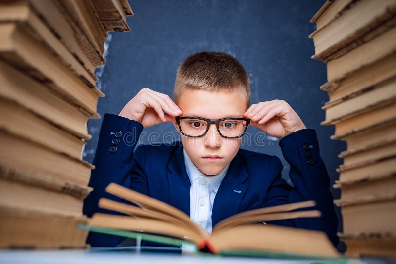Smart Boy in Glasses Sitting between Two Piles of Books and Read Book ...