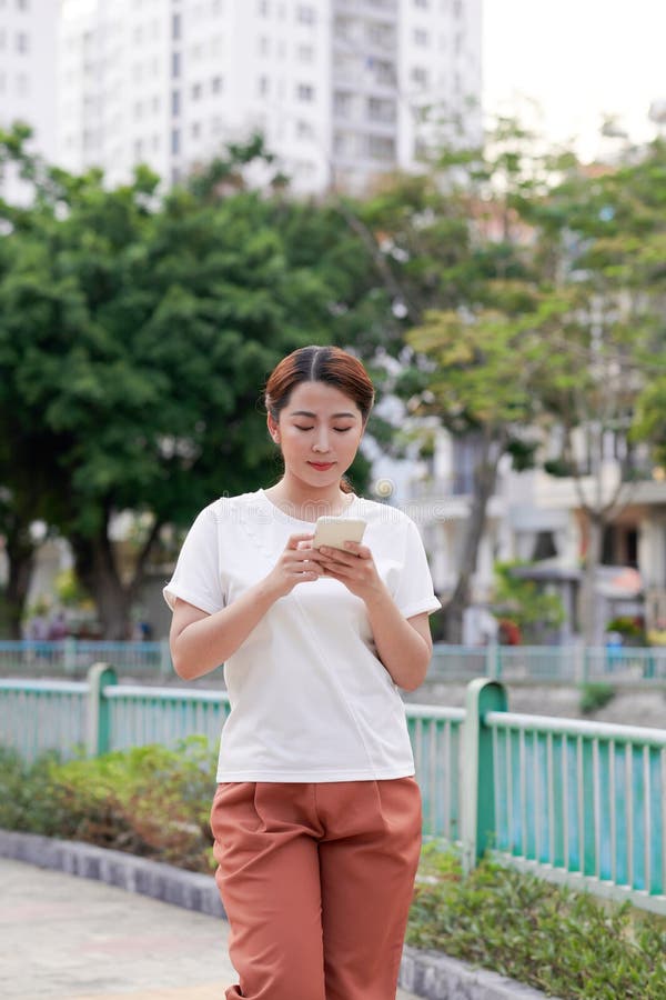 Smiling Attractive Young Woman Standing and Using Mobile Phone in Park ...