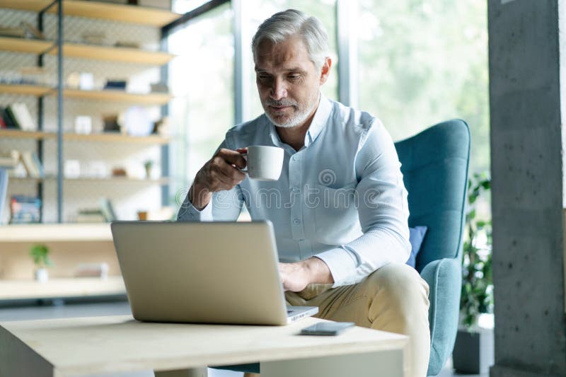 Smiling Businessman Using Laptop and Drinking Coffee while Working in ...