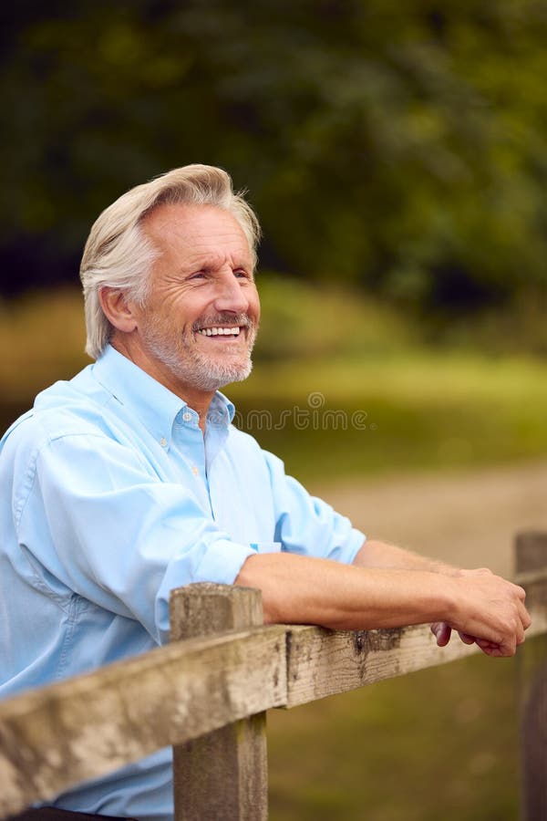 Smiling Casually Dressed Mature Or Senior Man Leaning On Fence On Walk