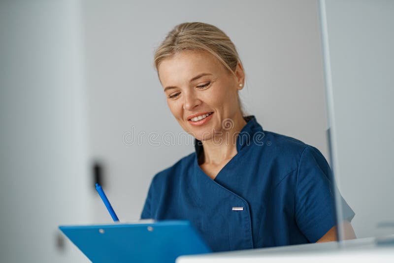 Smiling Doctor Making Notes in Clipboard Standing Near Reception in ... Smiling Doctor Making Notes in Clipboard Standing Near Reception in ...